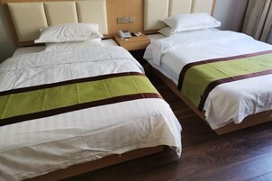 Quarto de hotel com duas camas, cada uma com roupa de cama branca e um cobertor riscado em verde e castanho. Há uma cabeceira de madeira, uma mesa de cabeceira com um telefone e uma luminária, e uma janela com cortinas.