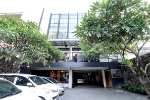Um edifício com estilo arquitetônico moderno, destacando uma fachada de vidro e um telhado plano. Vários carros estão estacionados em frente ao edifício, e uma árvore com folhas verdes é visível à esquerda.
