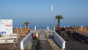 Um deck de madeira leva a uma área de praia com espreguiçadeiras e guarda-sóis, um mastro com bandeira e uma placa com informações de segurança.