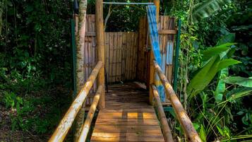 Uma ponte de madeira com guarda-corpo de bambu que leva a uma área coberta, cercada por vegetação densa.