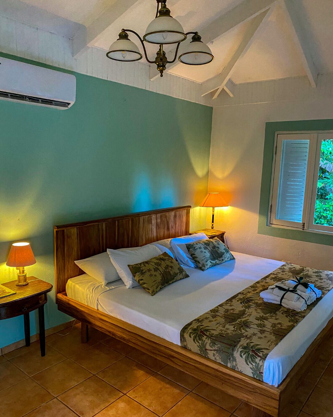 Quarto com cama de madeira, duas luminárias de cabeceira, janela com persianas e aparelho de ar condicionado.