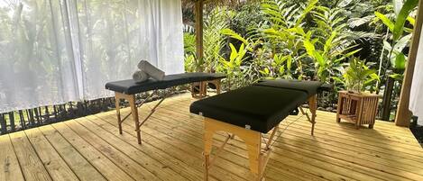 Um deck de madeira com duas mesas de massagem, um telhado de palha e plantas tropicais exuberantes ao fundo.