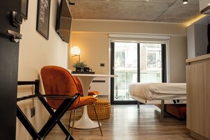 Quarto de hotel moderno com cama, escrivaninha, cadeira, planta em vaso e porta de vidro deslizante que dá para uma varanda.