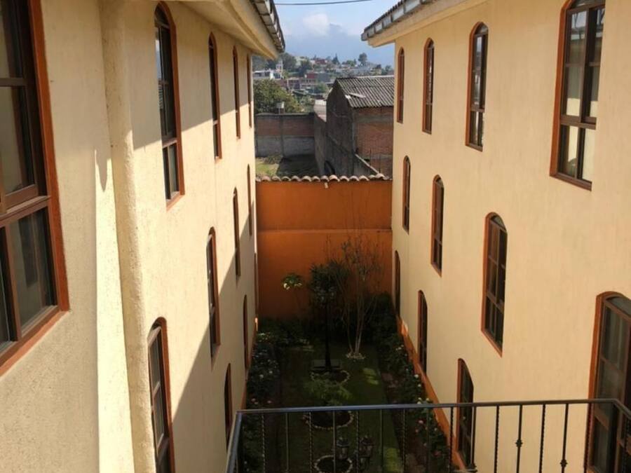 Um pátio estreito com um pequeno jardim, uma varanda com grades de metal e edifícios com janelas em arco.