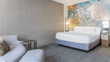 Quarto de hotel moderno com uma cama grande, um sofá, uma mesinha e uma mesa de cabeceira com abajur.