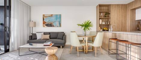 Uma sala de estar moderna com sofá, mesa de centro e mesa de jantar. Há uma área de cozinha com balcão e estantes.