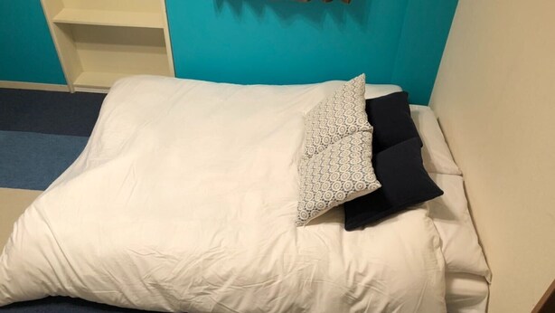 Cama com edredom branco e duas almofadas, uma estampada e outra preta lisa, em quarto com paredes verde-água e carpete azul.