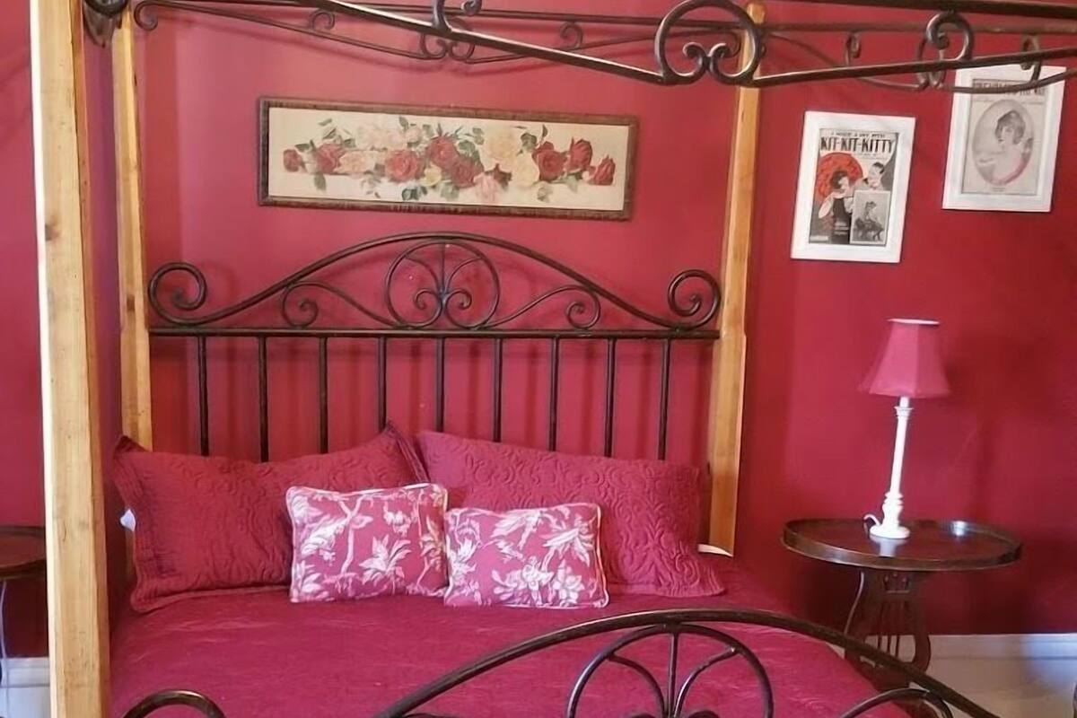 Uma cama de dossel com colcha e almofadas vermelhas, um candelabro e uma pequena mesa com abajur.