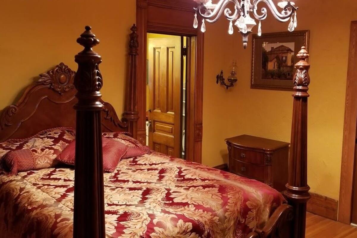 Uma cama com dossel com colcha florida, uma mesa de cabeceira de madeira, uma porta de madeira e um espelho na parede.