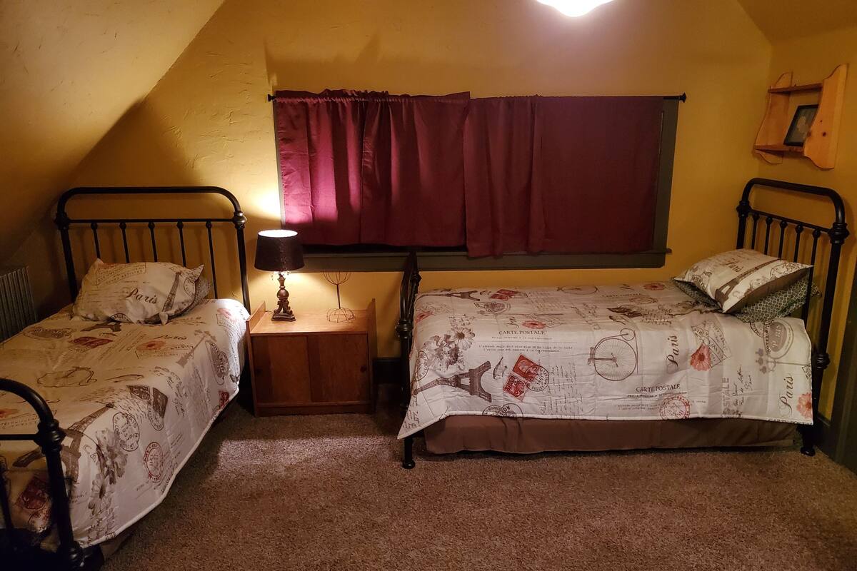 Quarto com duas camas de solteiro, mesa de cabeceira com abajur, janela com cortinas e prateleira fixada na parede.