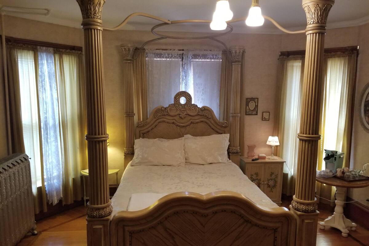 Uma cama com dossel e estrutura de madeira ornamentada na cabeceira e na peseira, ladeada por colunas altas. Uma mesa de cabeceira com abajur e um pequeno vaso está visível.