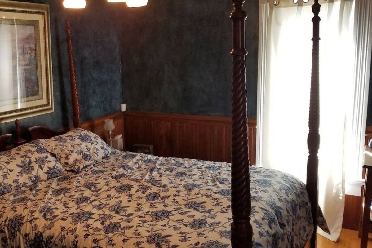 Uma cama com dossel e roupa de cama floral, um quadro na parede e uma janela com cortinas.