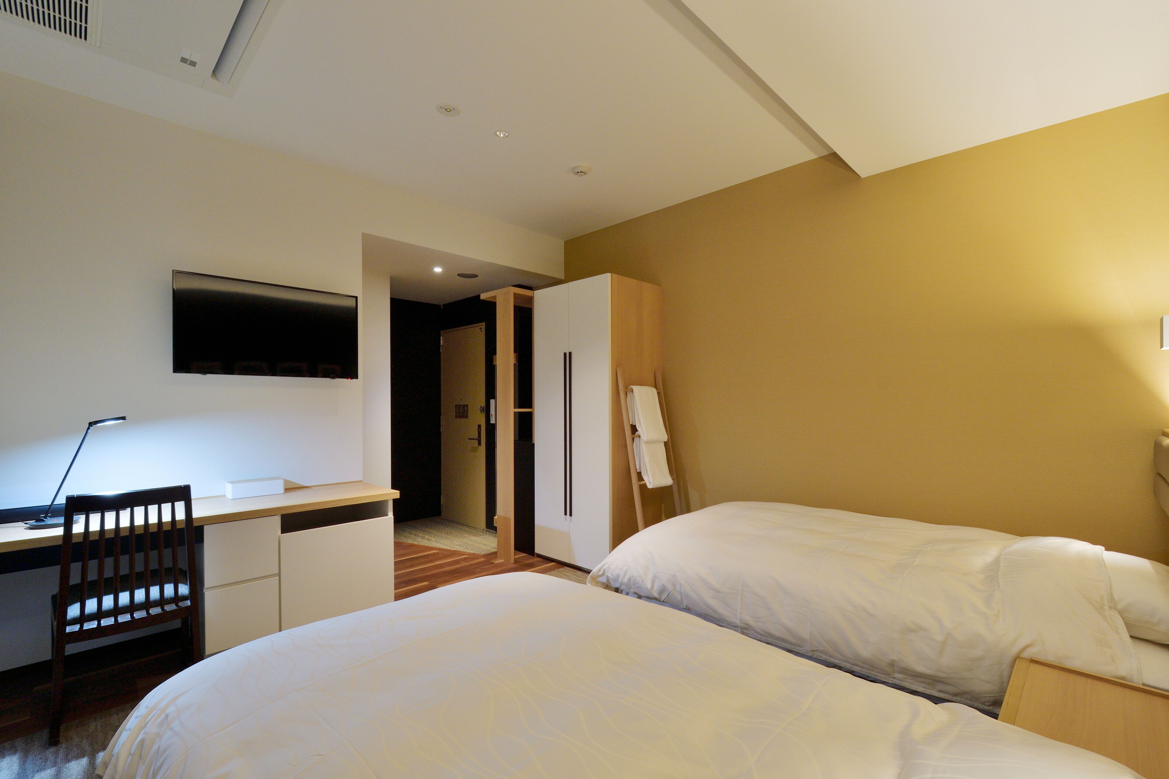 Quarto de hotel com duas camas, uma escrivaninha, uma cadeira, uma televisão e um guarda-roupa.