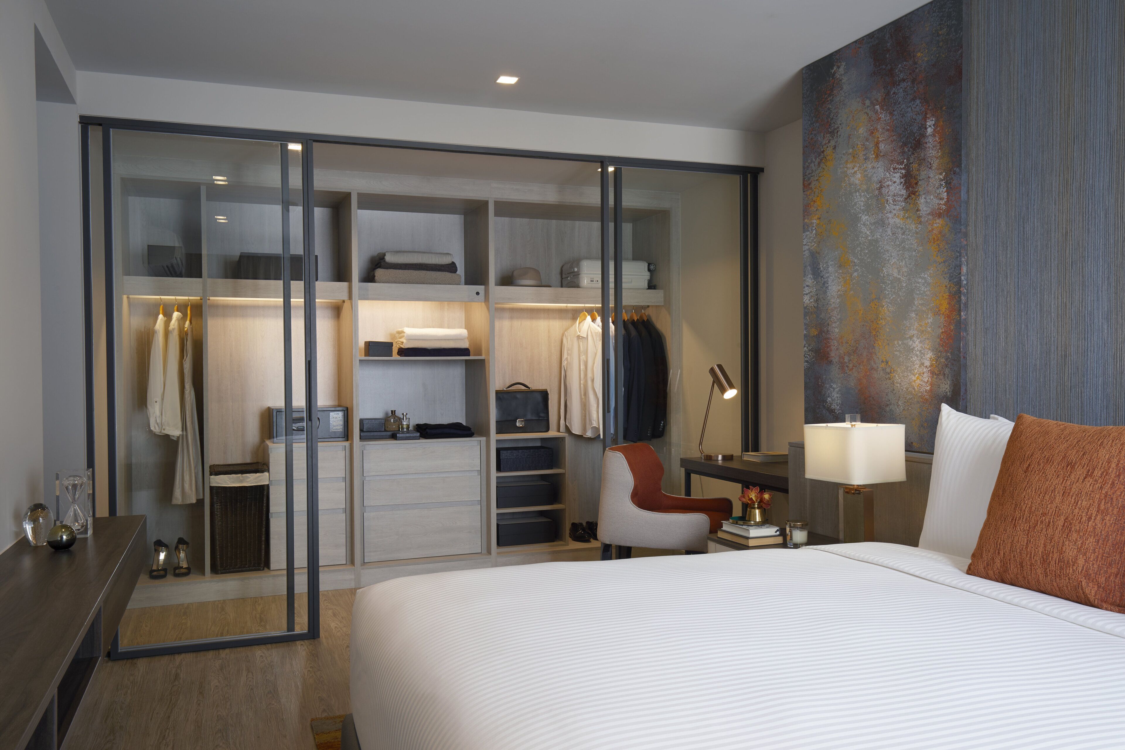 Quarto de hotel moderno com uma cama grande, uma mesa com luminária, uma cadeira, um guarda-roupa com portas de correr e um quadro na parede.
