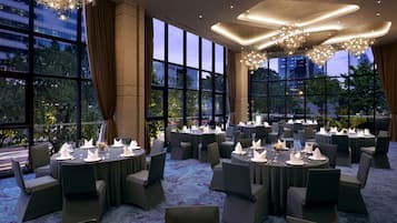 Uma ampla sala de jantar com mesas redondas preparadas para um evento formal, grandes janelas com vista para a cidade e luminárias modernas.