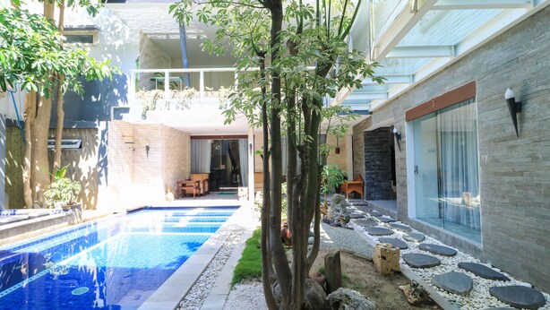 Uma área moderna de piscina ao ar livre, com piscina azul cristalina, rodeada por um caminho de pedras e uma árvore no centro.