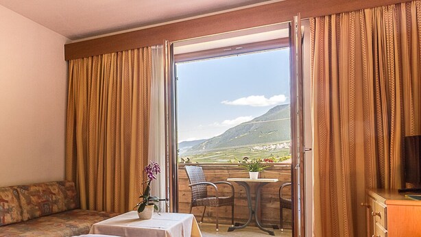 Quarto de hotel com cama, sofá, uma pequena mesa com uma planta e varanda com mesa e cadeiras.
