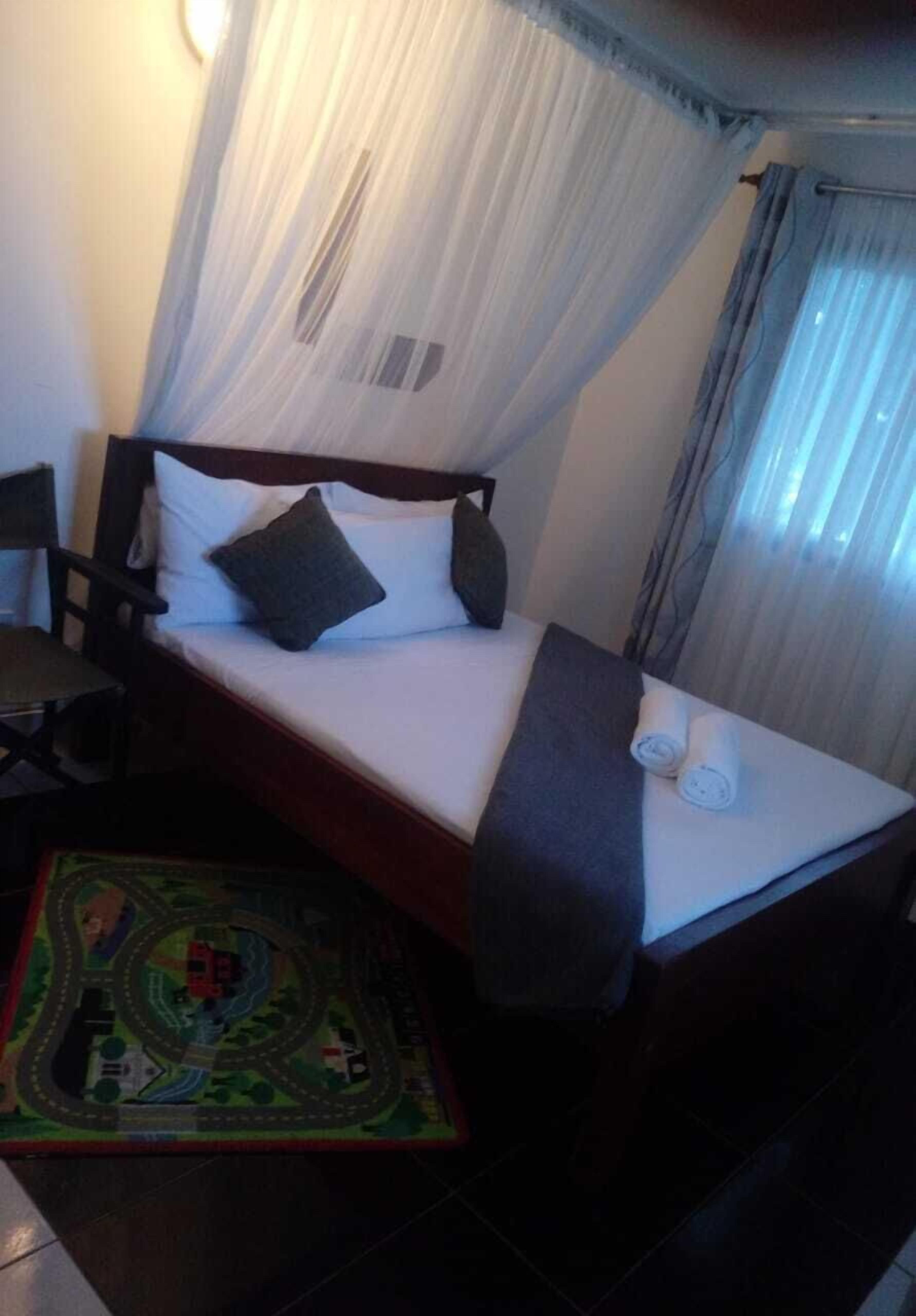 Uma cama de solteiro com cabeceira de madeira, roupa de cama branca e um cobertor grosso escuro. À esquerda, há uma pequena mesa com duas toalhas enroladas e uma cadeira.