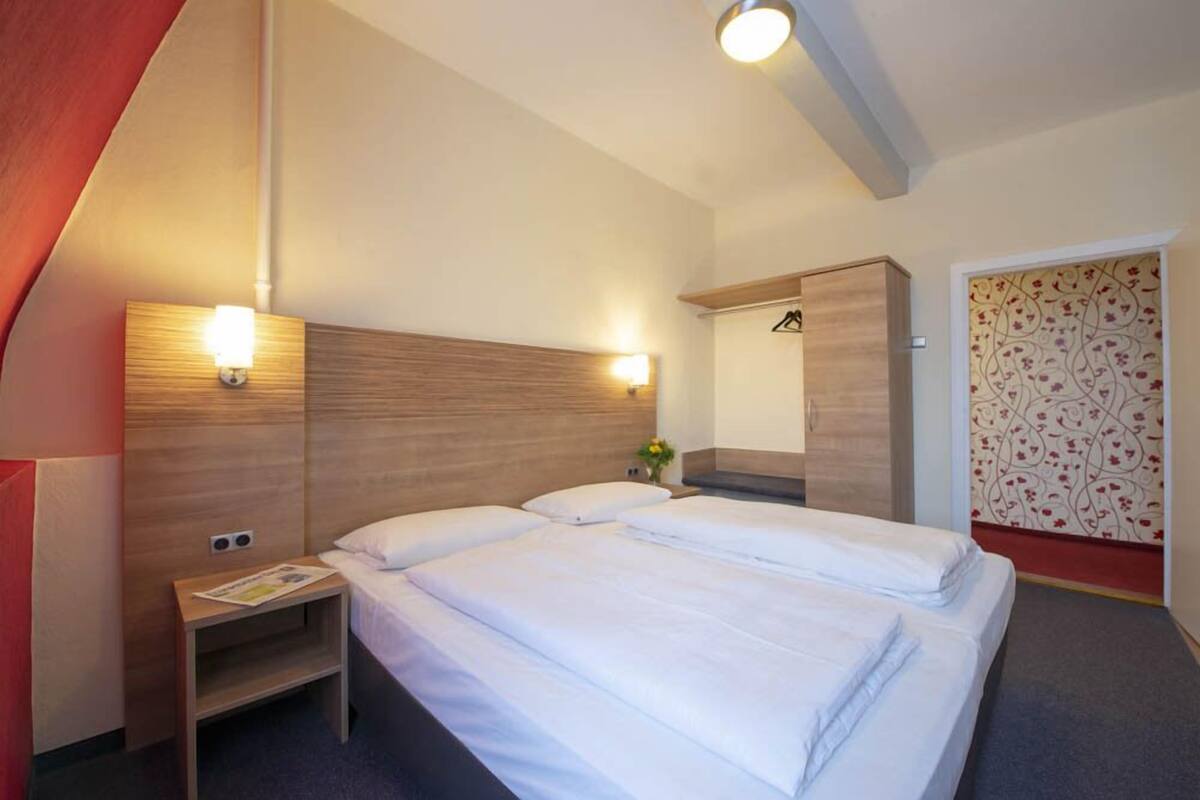 Quarto de hotel com cama de casal, luminárias de cabeceira, cabeceira de madeira, mesa de cabeceira e papel de parede com padrão floral.