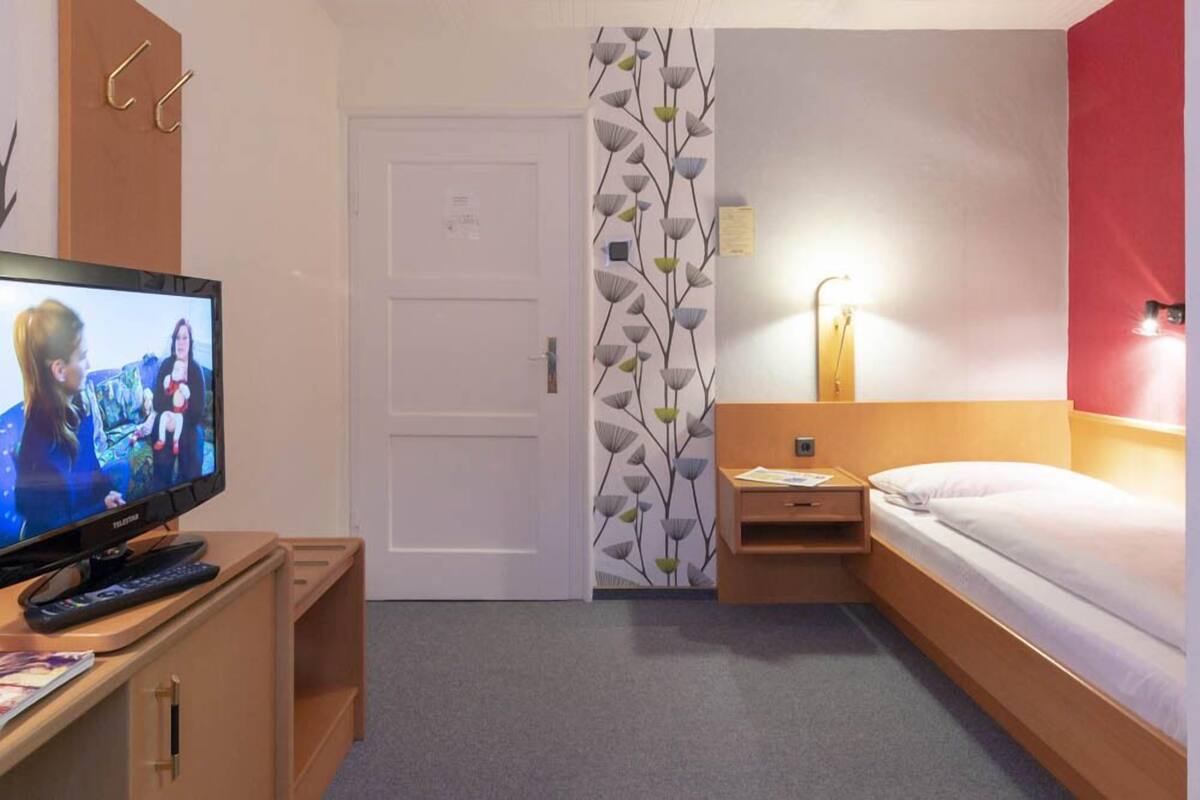Quarto de hotel com televisão sobre uma escrivaninha de madeira, uma cama com colchão branco, uma mesa de cabeceira com abajur e uma porta com papel de parede estampado com folhas.