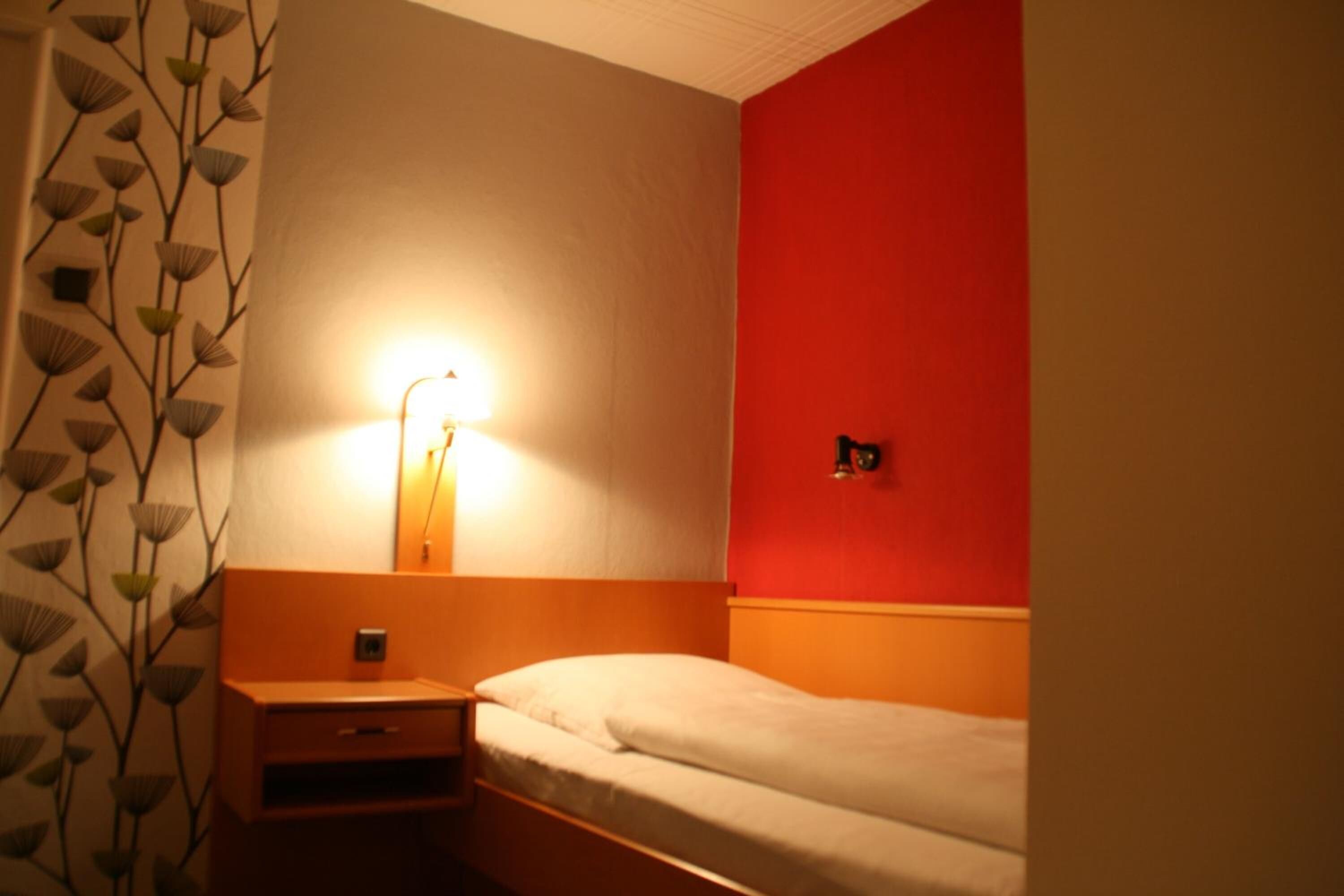 Quarto com uma cama de solteiro, mesa de cabeceira, luminária na parede e uma parede com detalhes vermelhos.