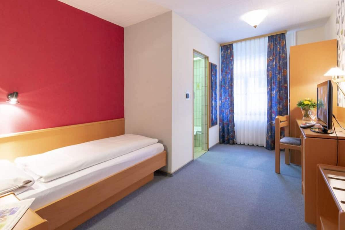 Quarto de hotel com uma cama de solteiro, uma mesa com cadeira, televisão e janela com cortinas.