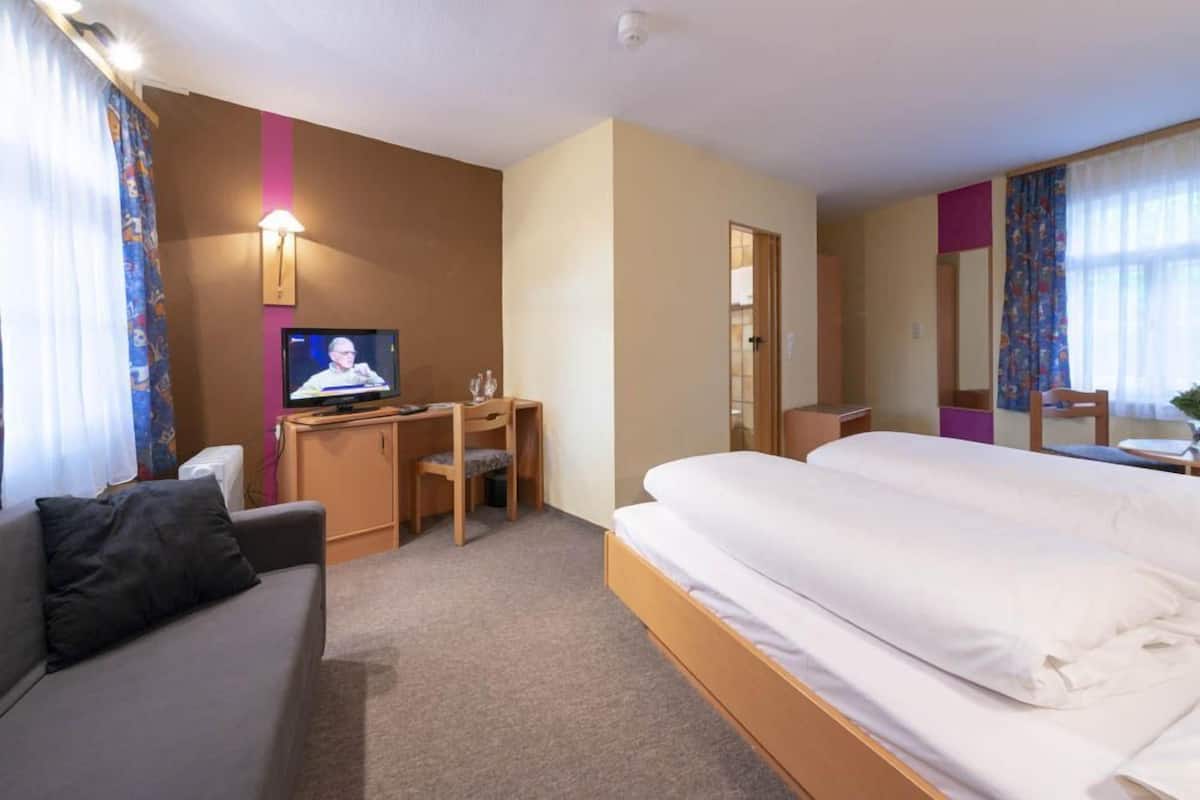 Quarto de hotel com cama, escrivaninha com computador, cadeira, sofá e televisão.