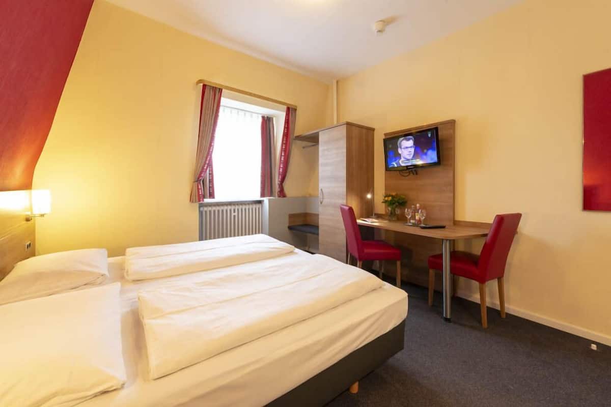 Quarto de hotel com cama, escrivaninha, cadeira, televisão e janela com cortinas.
