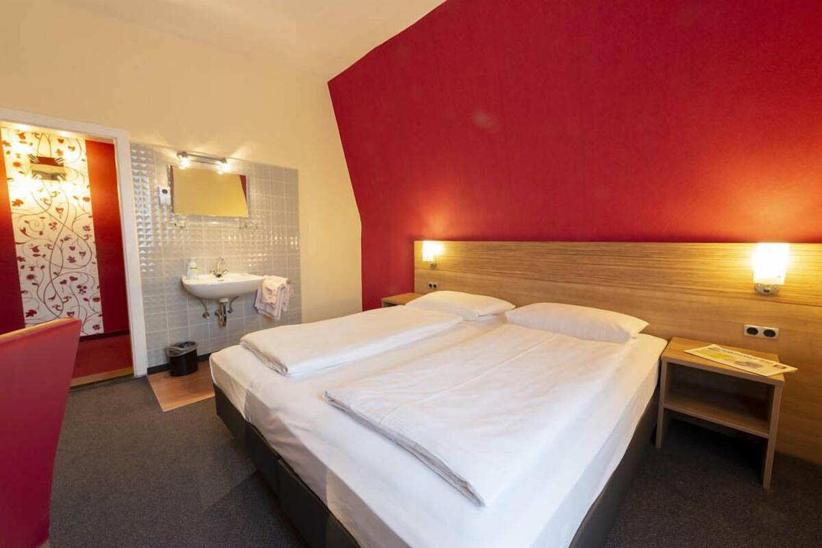 Quarto de hotel com uma cama grande, uma mesa de cabeceira, um armário e um banheiro com pia e espelho, e uma parede com detalhes vermelhos.