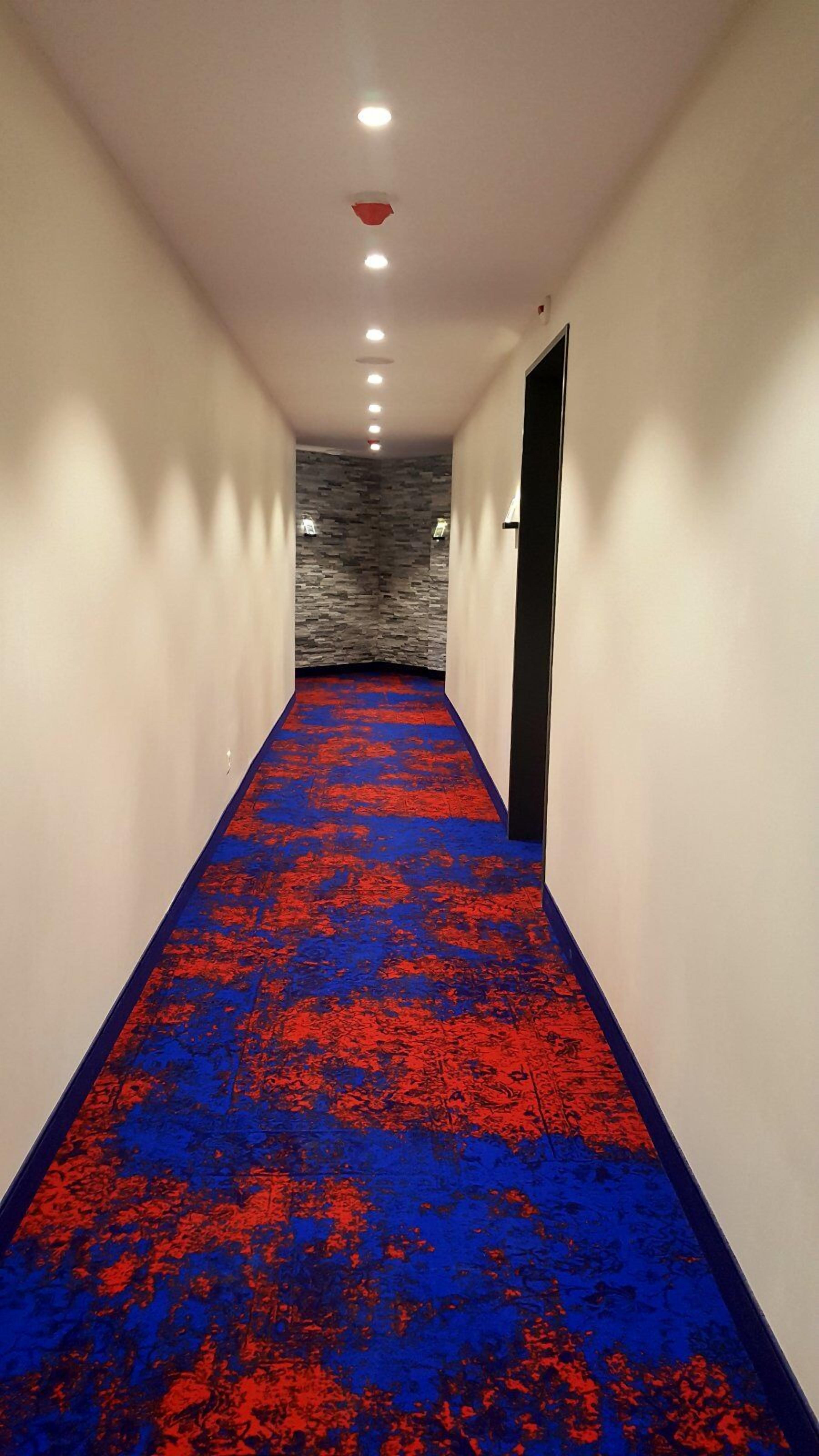 Um corredor com carpete vermelho e azul, paredes brancas e uma parede de pedra no final.