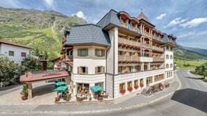 Um hotel com vista para as montanhas, área externa com mesas e um letreiro que diz "Alpenhof".