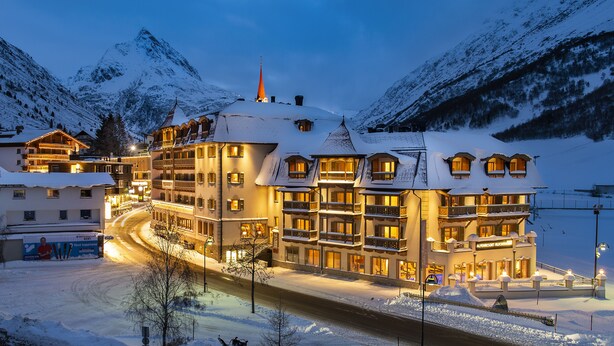 Hotel coberto de neve com várias varandas e um teleférico ao fundo.