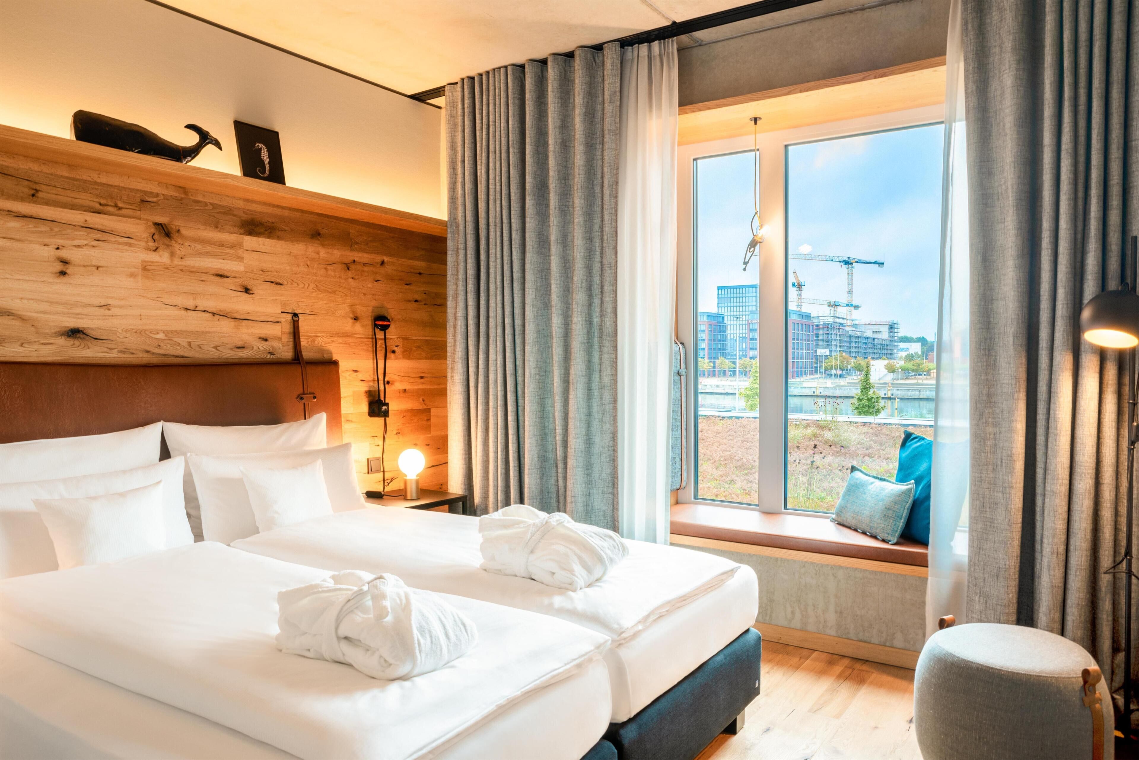 Quarto de hotel com uma cama grande, cabeceira de madeira, janela com vista para um canteiro de obras e um banco junto à janela.