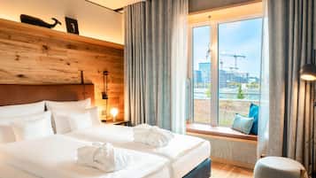 Quarto de hotel com uma cama grande, cabeceira de madeira, janela com vista para um canteiro de obras e um banco junto à janela.