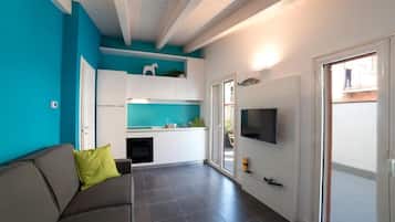 Uma sala de estar moderna com um sofá, televisão na parede e uma área de cozinha com armários e bancada.