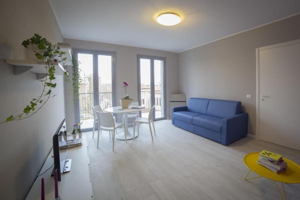 Uma sala de estar moderna com um sofá azul, uma mesa de jantar branca com cadeiras, uma mesinha lateral amarela e uma varanda com vista.