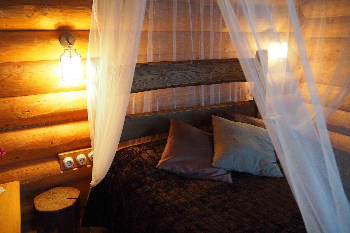 Um quarto aconchegante em estilo cabana, com cabeceira de madeira, cama com travesseiros, um banquinho pequeno e luminária fixada na parede.