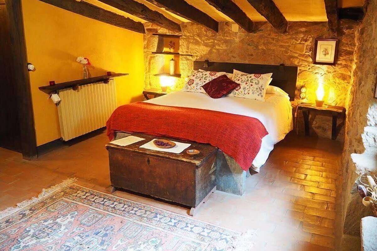 Quarto tradicional com parede de pedra, vigas de madeira, cama com manta vermelha e mesa de cabeceira com abajur.