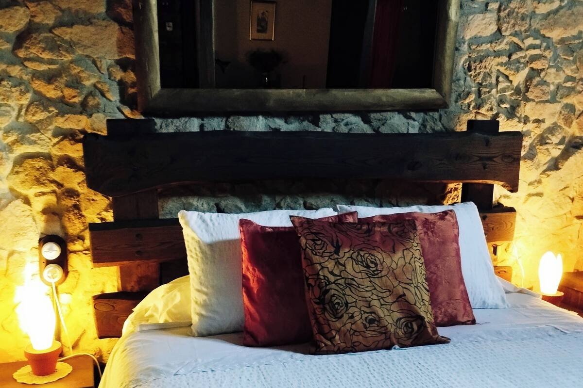 Uma cama com parede de pedra ao fundo, cabeceira de madeira e mesa de cabeceira com luminária.