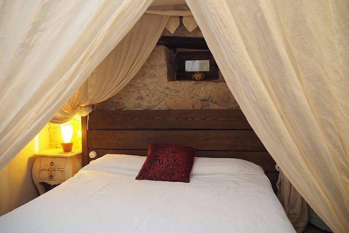 Quarto com cama, mesa de cabeceira, abajur e parede de pedra.