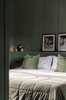 Cama bem arrumada com cobertura acolchoada, travesseiros verdes e uma estante com livros e uma lupa.
