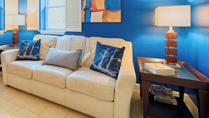 Uma sala de estar com sofá bege, parede azul, janela com persianas, luminária e uma pequena mesa com livros.
