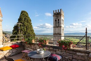 Varanda de pedra com uma mesa, cadeiras e plantas em vasos. Uma torre alta com janelas em arco visível ao fundo.