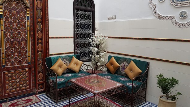 Um interior tradicional marroquino com um sofá estampado, uma mesa pequena e elementos decorativos como uma janela com detalhe em metal e uma parede com desenhos geométricos.