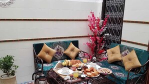 Uma área de estar acolhedora ao ar livre, com piso xadrez, uma mesa de metal posta para o café da manhã e um vaso de planta.