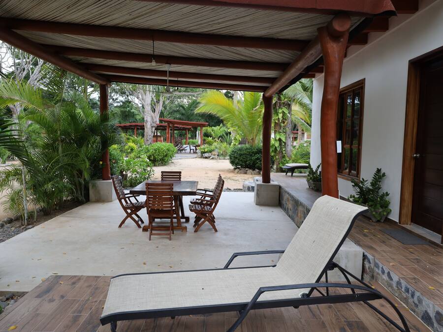 Um pátio externo com mesa e cadeiras, rodeado por plantas e uma pérgula de madeira.
