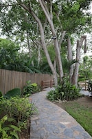Um caminho de pedra serpenteia por um jardim exuberante, com árvores grandes e diversas plantas.