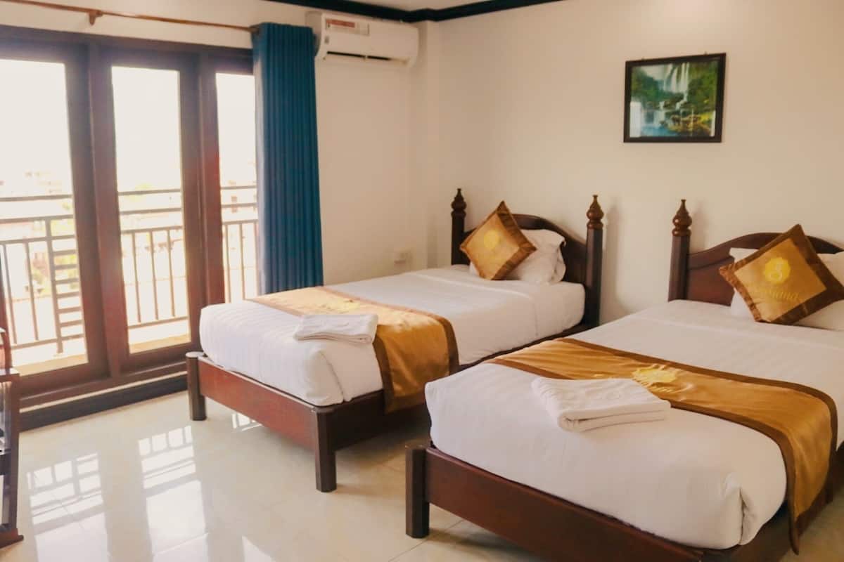 Duas camas de casal com estruturas de madeira e roupa de cama branca, um quadro na parede e vista através de uma porta de vidro.