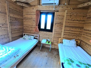 Uma cabana pequena com duas beliches de madeira, uma janela com cortinas, uma mesinha com uma cadeira e um ar condicionado.