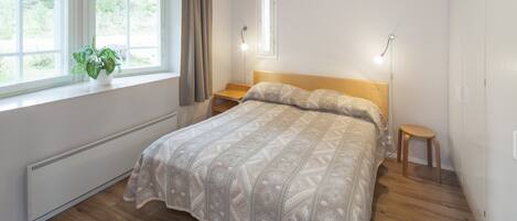 Quarto com cama, janela, radiador, mesa de cabeceira e planta em vaso.